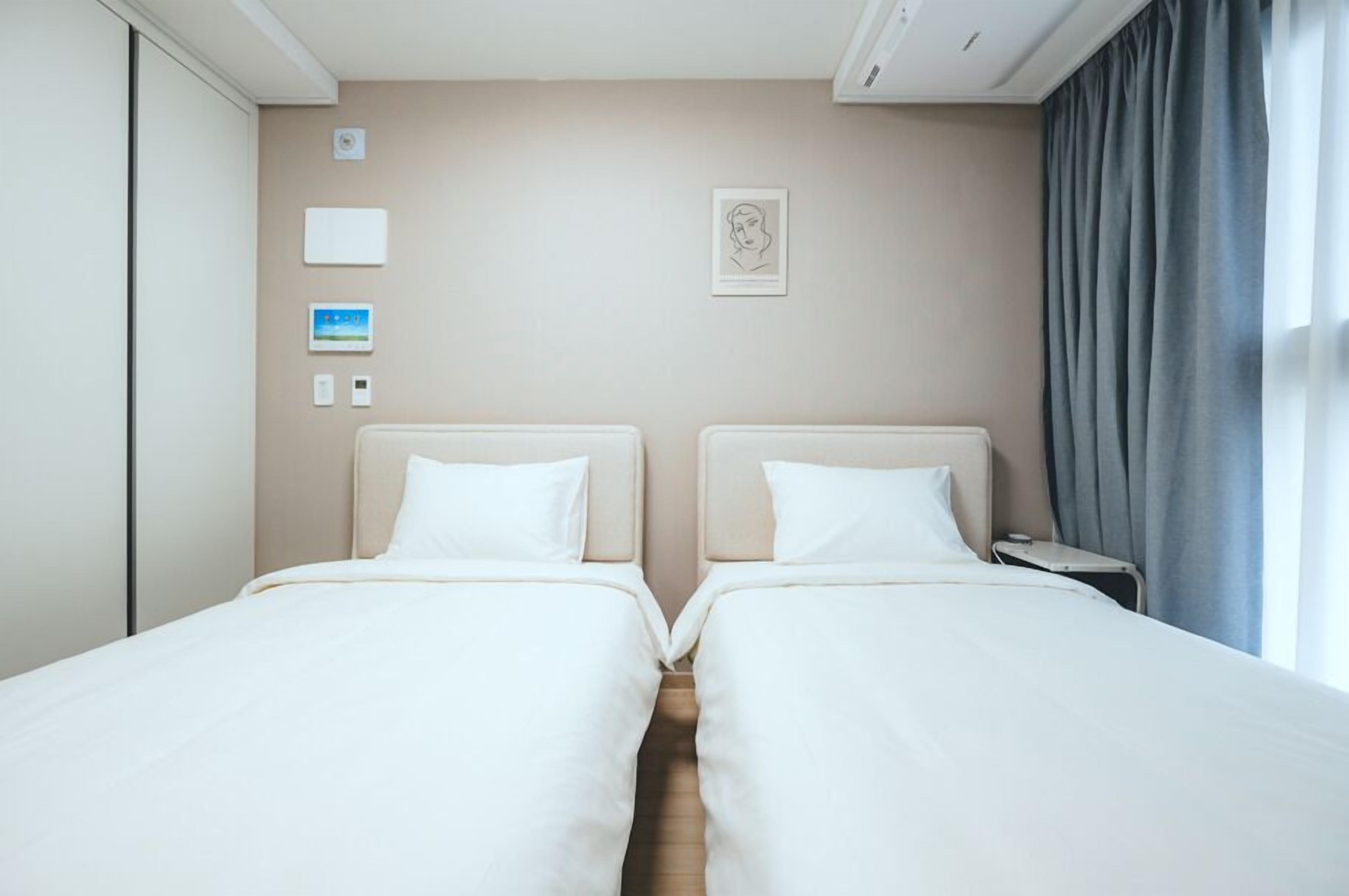 Quarto de hotel com duas camas, uma mesa de cabeceira, termostato na parede e uma janela com cortinas.