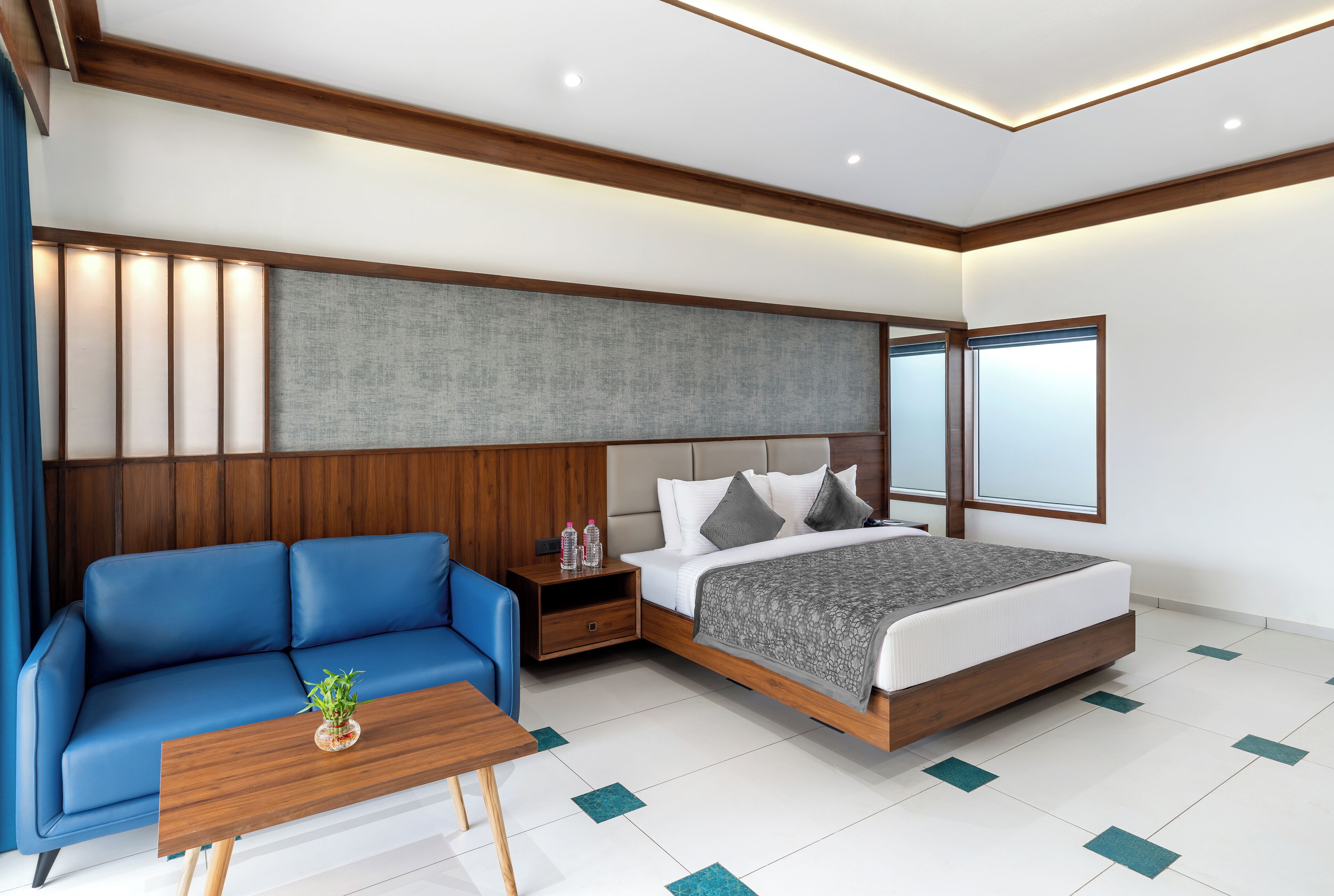 Quarto moderno com uma cama, um sofá azul, uma mesinha de cabeceira de madeira e uma janela grande.