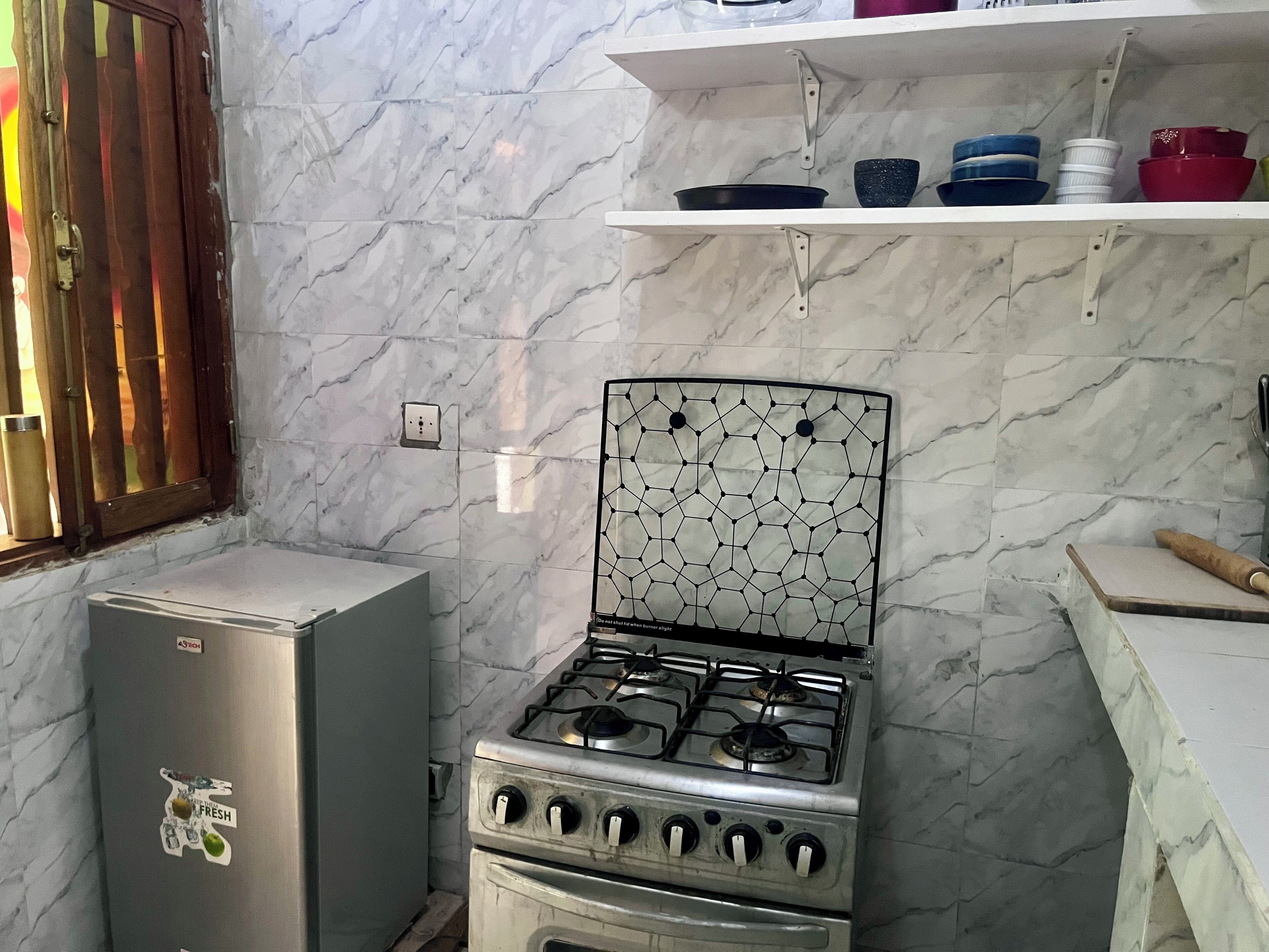 Uma cozinha com fogão a gás, uma geladeira pequena e uma janela com persiana de madeira.