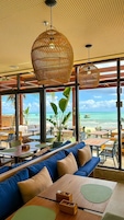 Um restaurante à beira-mar com luminárias de vime pendentes, assentos acolchoados em azul e amplas janelas com vista para o oceano.