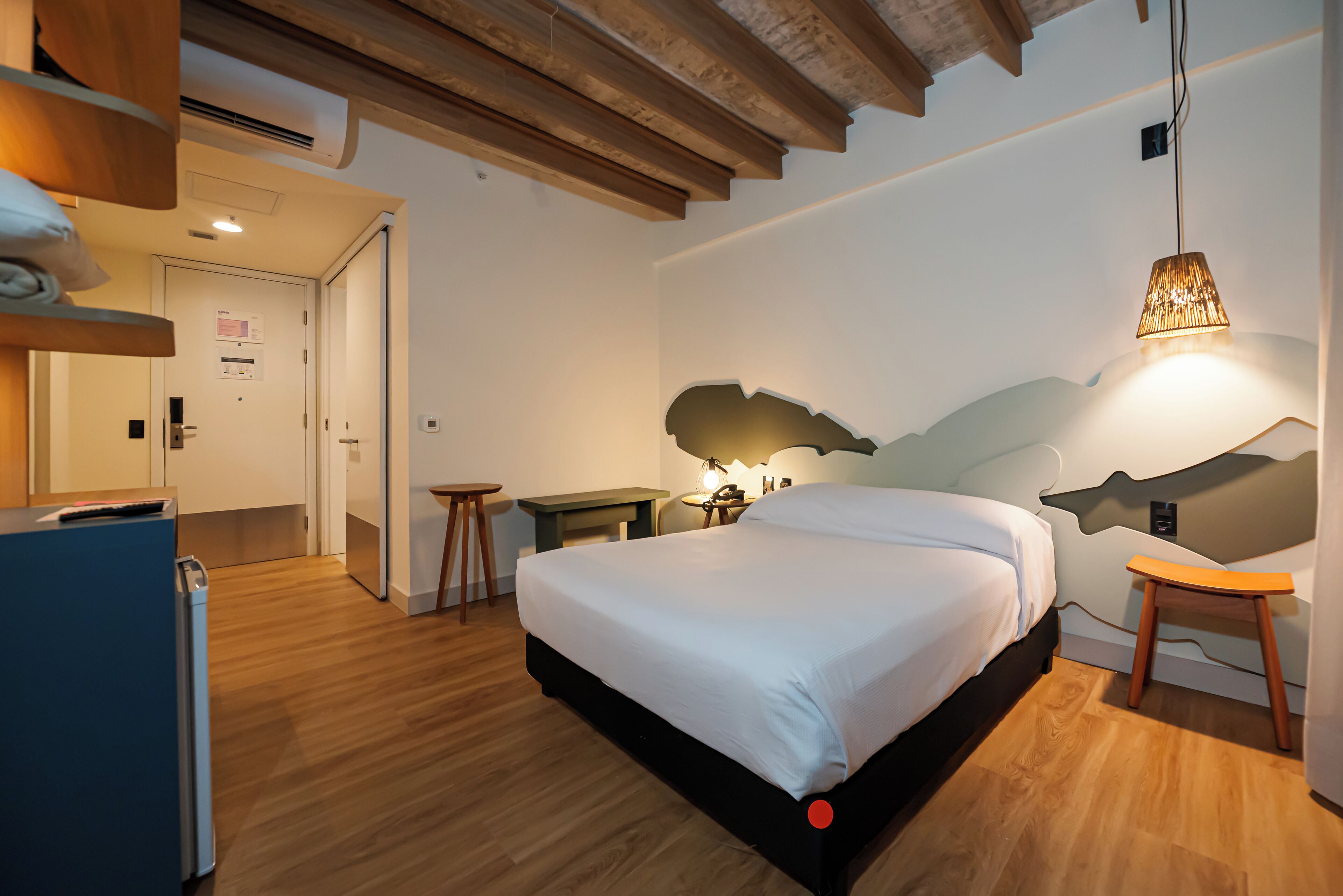 Quarto moderno com cama, mesa de cabeceira, abajur e um banquinho pequeno.