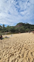 Uma praia arenosa com pessoas tomando sol e nadando, rodeada por vegetação exuberante e uma colina ao fundo.