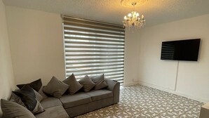 Uma sala de estar com sofá, piso estampado, um lustre e uma televisão fixada na parede.