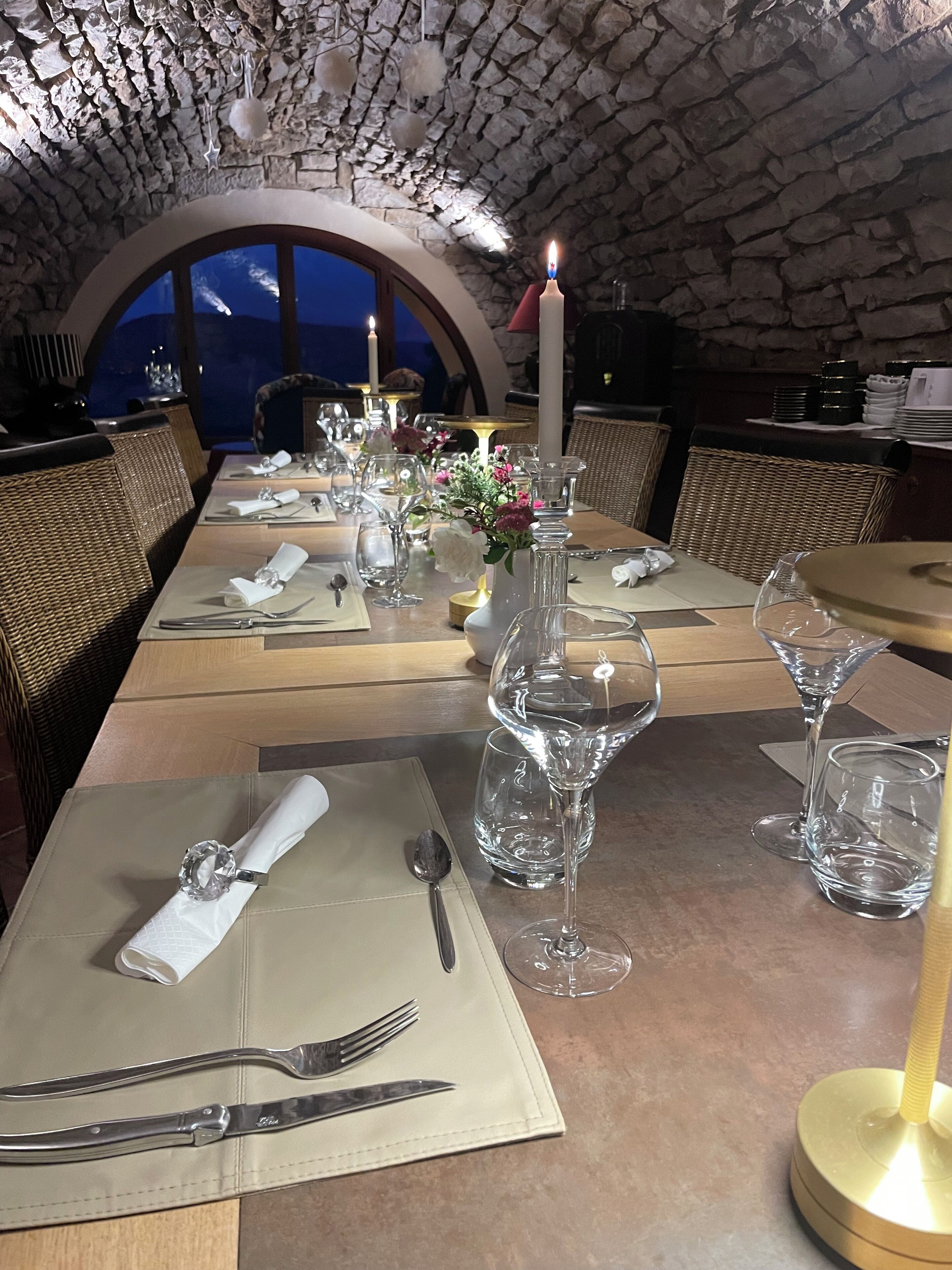 Restaurante com teto de pedra e uma grande janela em arco. Mesas postas com copos de vinho, pratos e talheres.