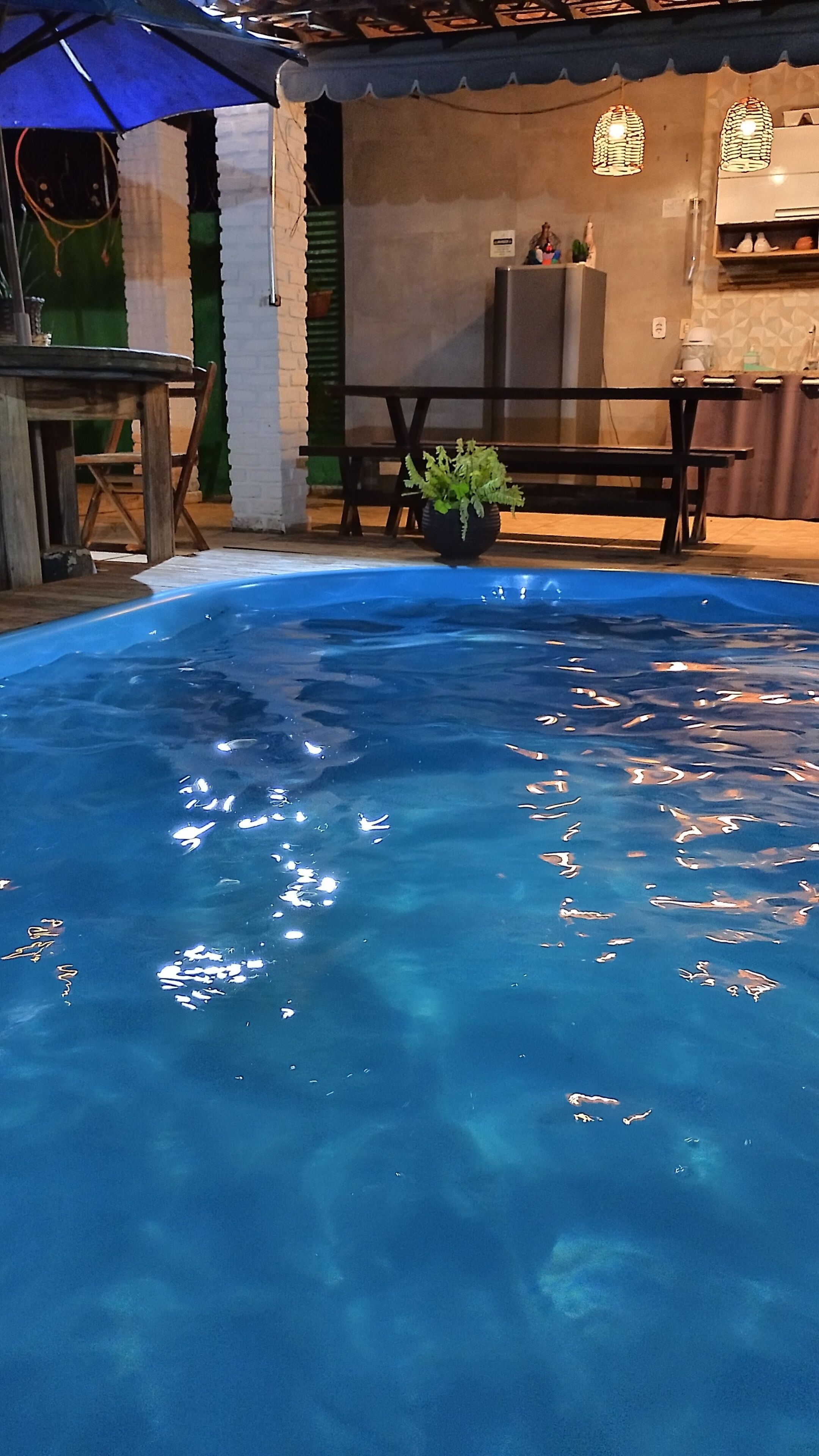 Piscina com água cristalina, um banco de madeira, um vaso de planta e uma mesa com cadeiras.