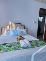 Uma cama bem arrumada com lençóis brancos e um edredom com padrão de folhas tropicais. Há toalhas enroladas e um pequeno buquê de flores sobre a cama.