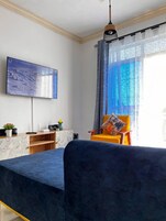 Uma sala de estar moderna com uma grande televisão de tela plana na parede, um sofá confortável, uma pequena mesa de madeira com um vaso e uma planta, e uma janela com cortinas azuis.
