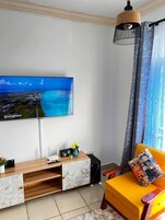 Uma sala de estar com televisão na parede, um móvel de entretenimento de madeira, um sofá amarelo e uma almofada decorativa estampada.