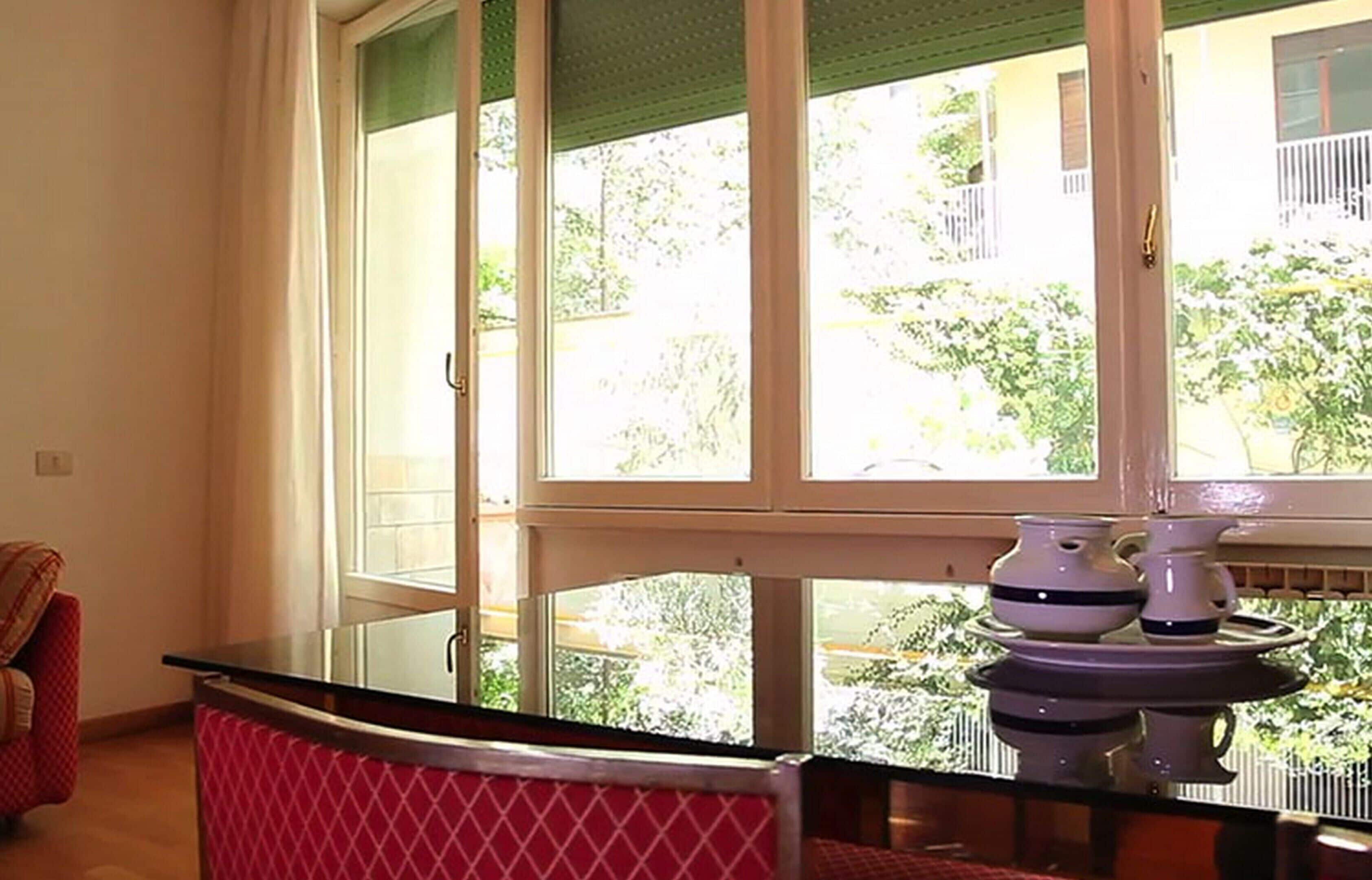 Quarto com uma grande janela, um banco estofado vermelho e uma mesa de vidro com objetos decorativos.