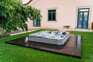Uma banheira de hidromassagem em um quintal com um deck de madeira, um caminho de pedra e uma casa com fachada rosa e portas azuis.