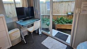 Quarto compacto com escrivaninha, cadeira e televisão. Possui porta de vidro deslizante para área externa com cerca de madeira e plantas.