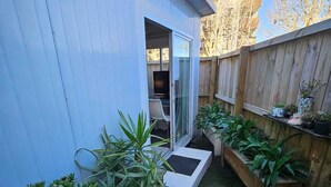 Um pequeno quintal cercado com uma porta de correr de vidro que dá para um espaço de pátio. Há canteiros elevados com diversas plantas e uma pequena mesa branca.