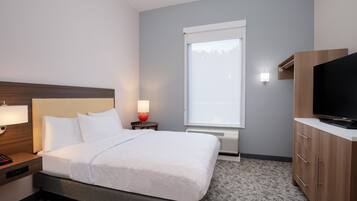 Quarto de hotel com cama, mesa de cabeceira, luminária, televisão e janela com persianas.