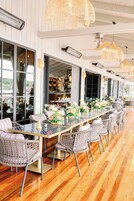Uma longa mesa de jantar com cadeiras de vime e centros de mesa florais, em um ambiente com grandes janelas e luminárias pendentes.