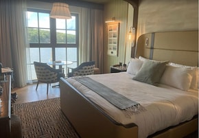 Quarto de hotel com uma cama grande, uma mesa, uma cadeira e vista para a água pela janela.