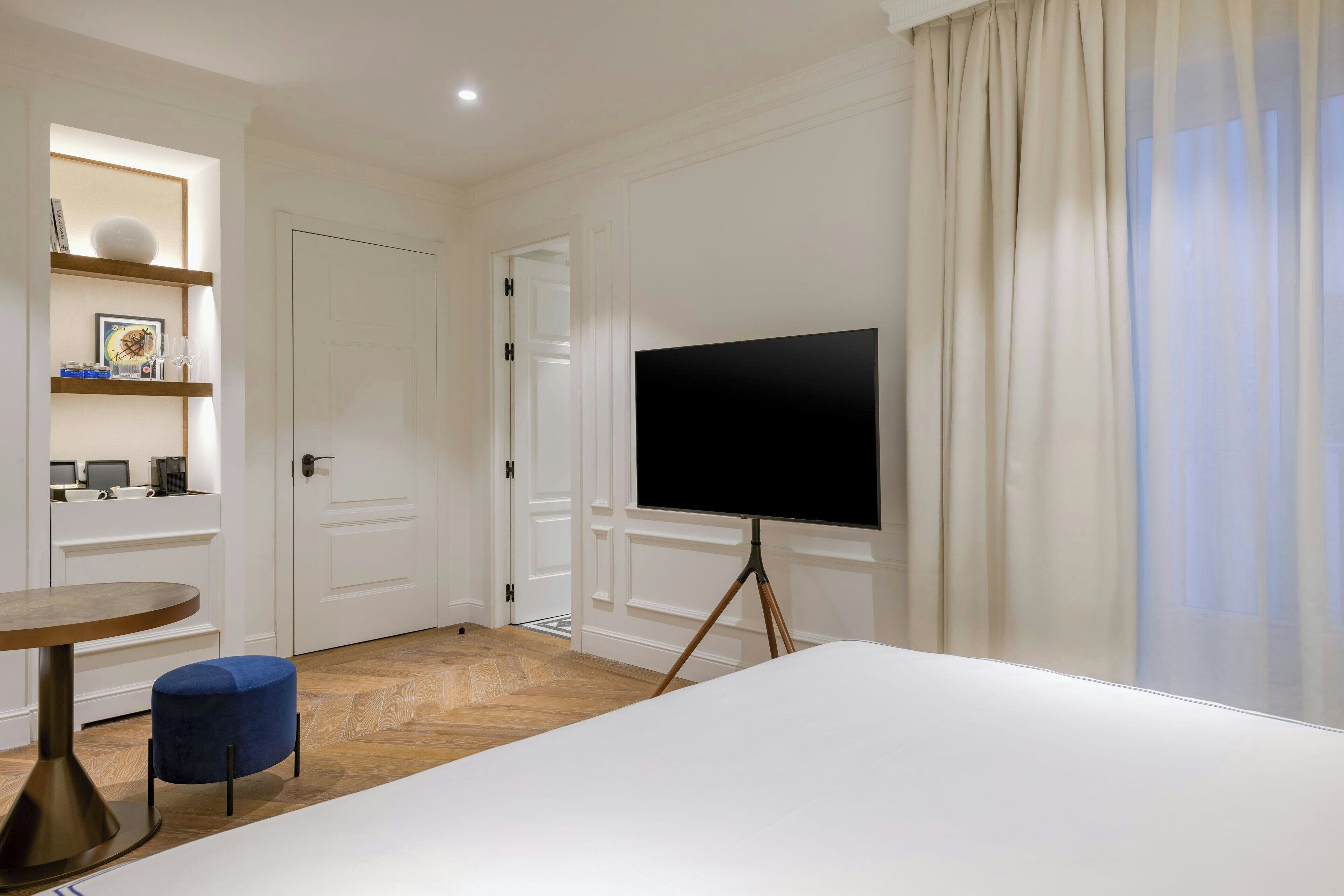 Quarto de hotel com uma cama grande, uma televisão de tela plana em um suporte de tripé, uma mesinha redonda com um banquinho azul e um guarda-roupa branco.