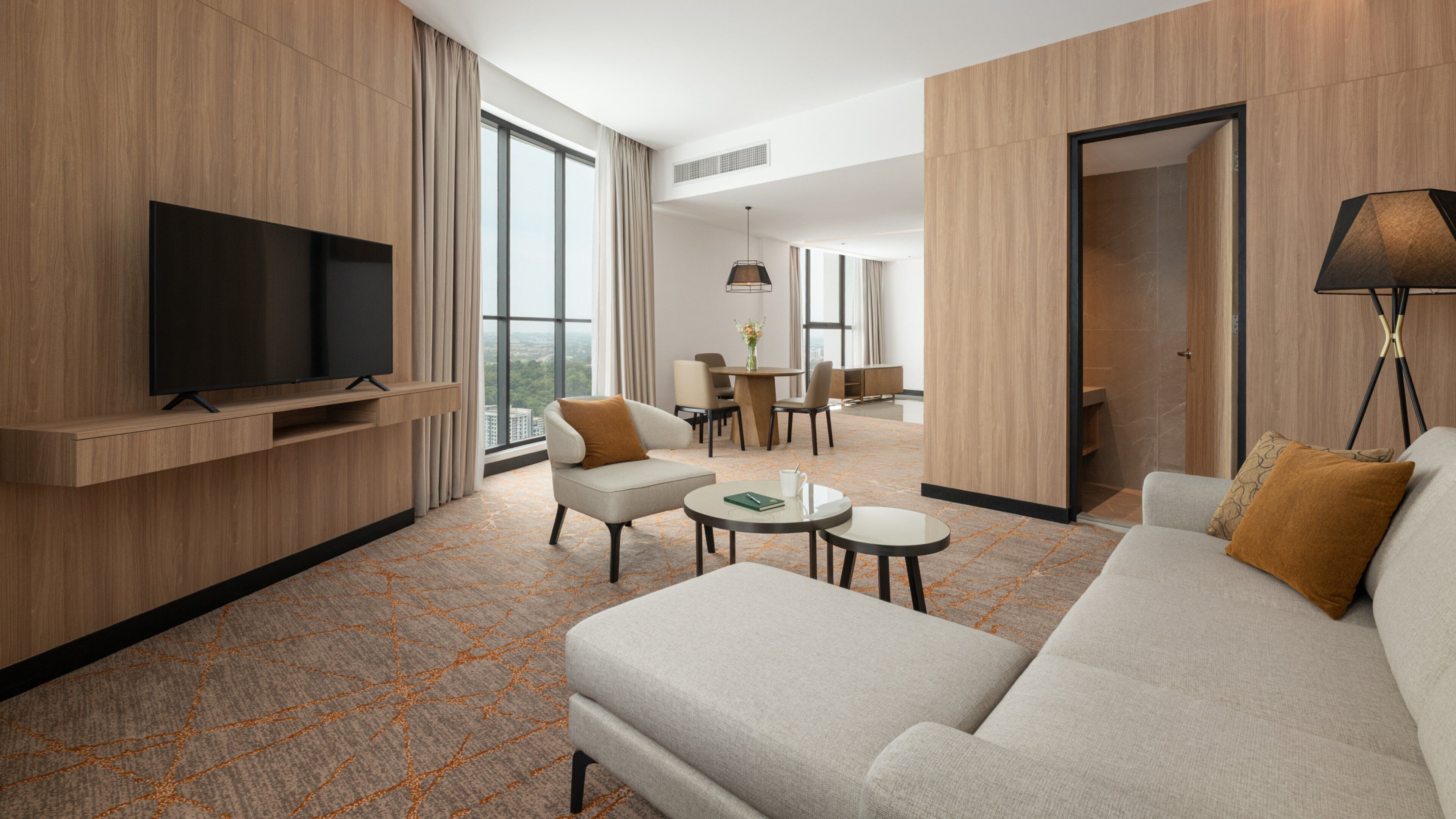 Quarto de hotel moderno com televisão de tela plana, sofá, duas poltronas, mesa de jantar com cadeiras e uma grande janela com vista para a cidade.