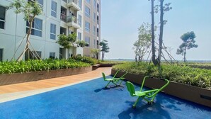 Terraço no telhado com piso azul, cadeiras verdes e vista para um prédio e árvores.
