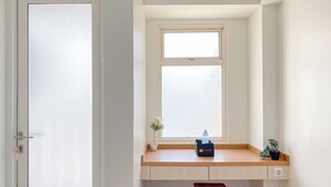 Um quarto pequeno e luminoso, com uma mesa, uma janela, um banquinho e uma porta.