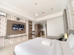 Quarto de hotel com cama grande, televisão na parede, pequena cozinha e banheiro visível através de uma porta aberta.