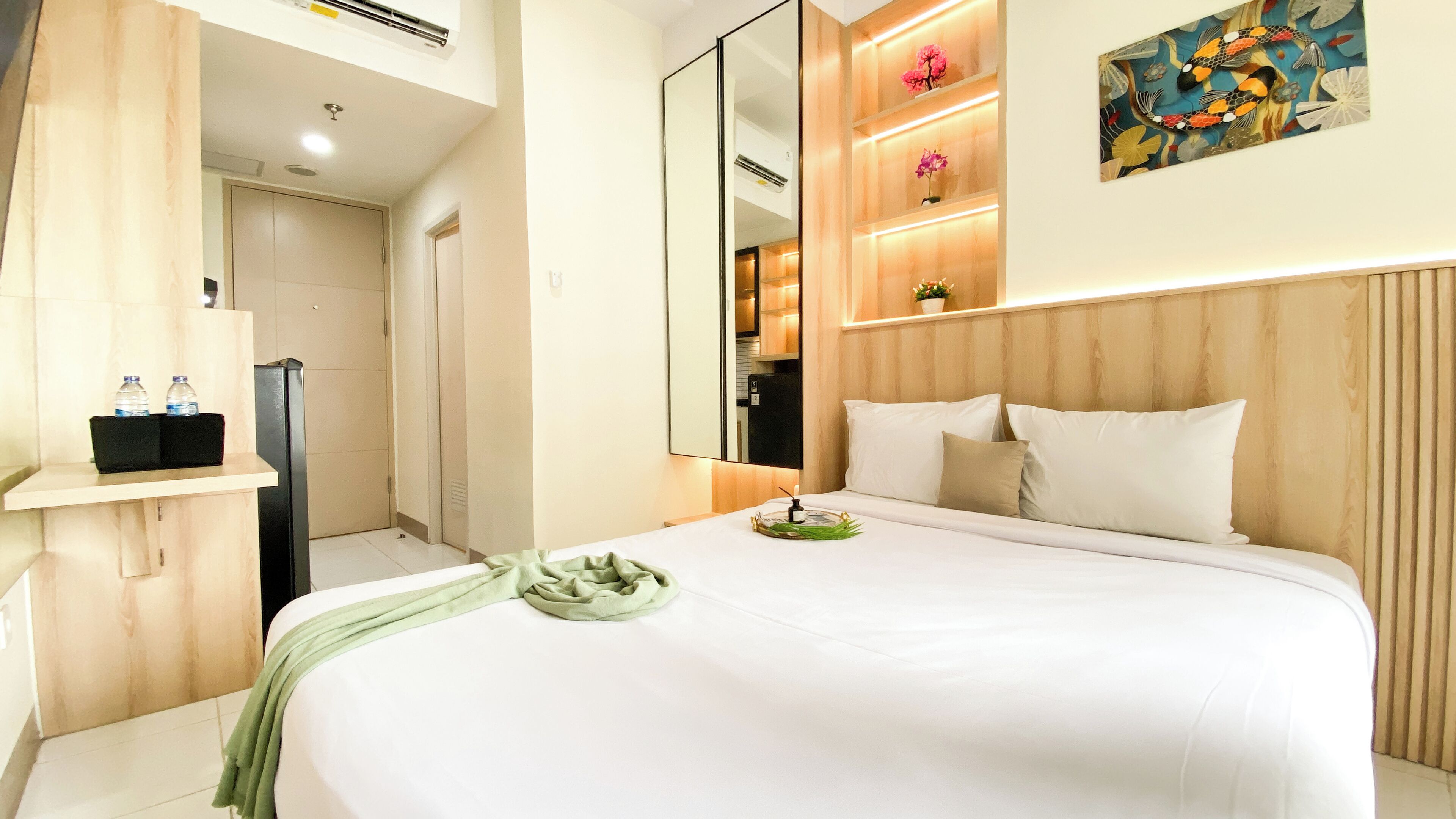 Quarto de hotel com uma cama grande, um espelho, uma pequena prateleira com garrafas de água e um quadro na parede.