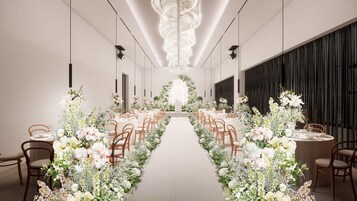 Um salão amplo com uma passagem central que leva a um arco floral, rodeado por mesas e cadeiras.