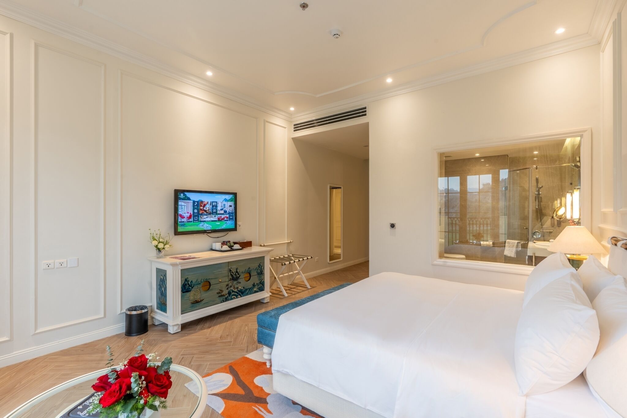Quarto de hotel com cama, televisão, mesa de apoio, espelho e banheiro visível através de uma porta de vidro.