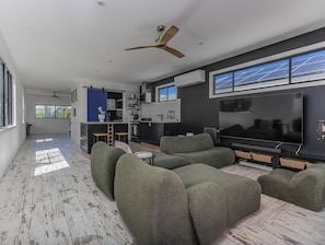 Uma sala de estar moderna com uma televisão de tela plana grande, sofás estofados em verde, um ventilador de teto e vista para painéis solares lá fora.