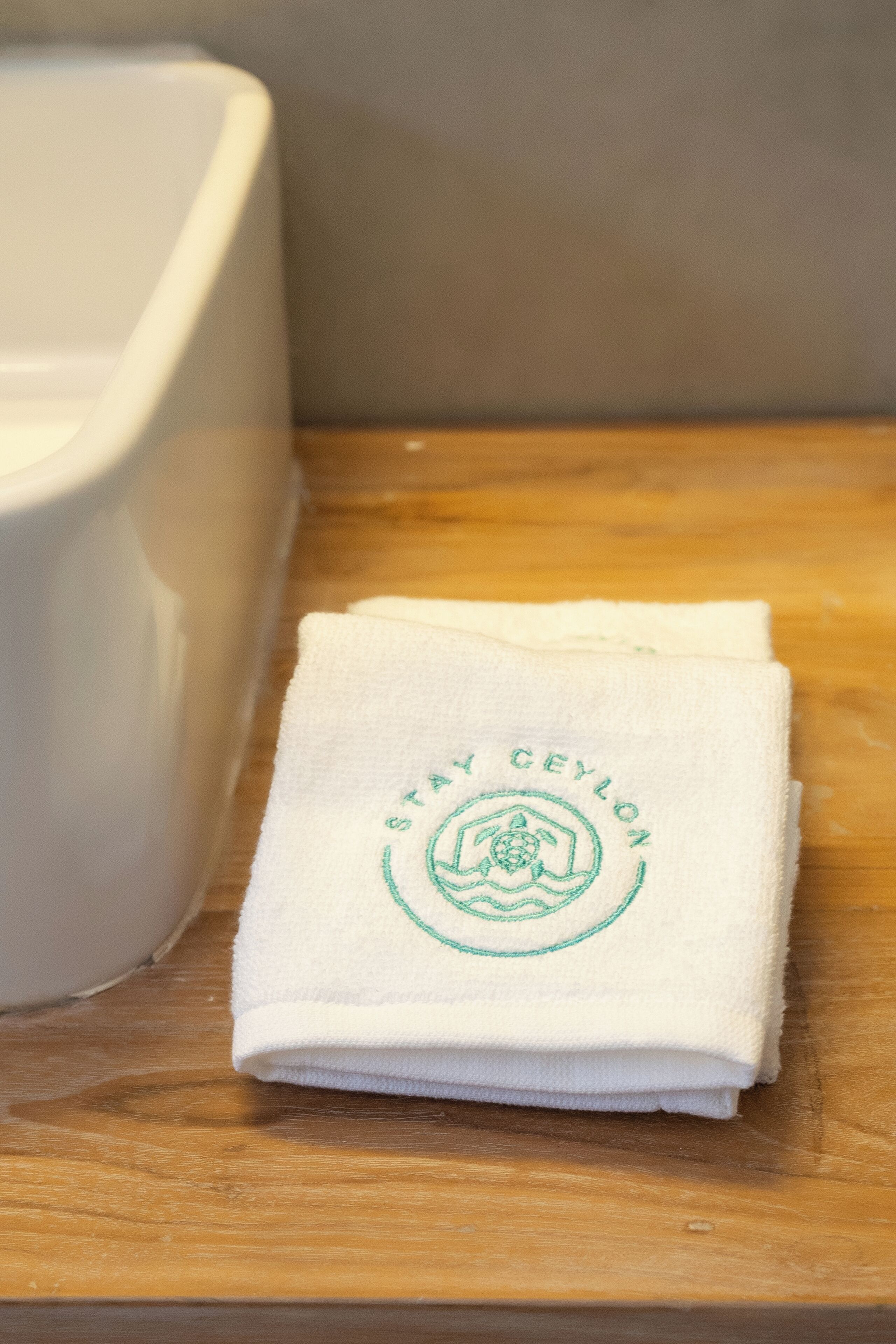 Toalha branca dobrada com a inscrição "STA. GEYLON" bordada, colocada sobre uma superfície de madeira ao lado de um vaso sanitário branco.