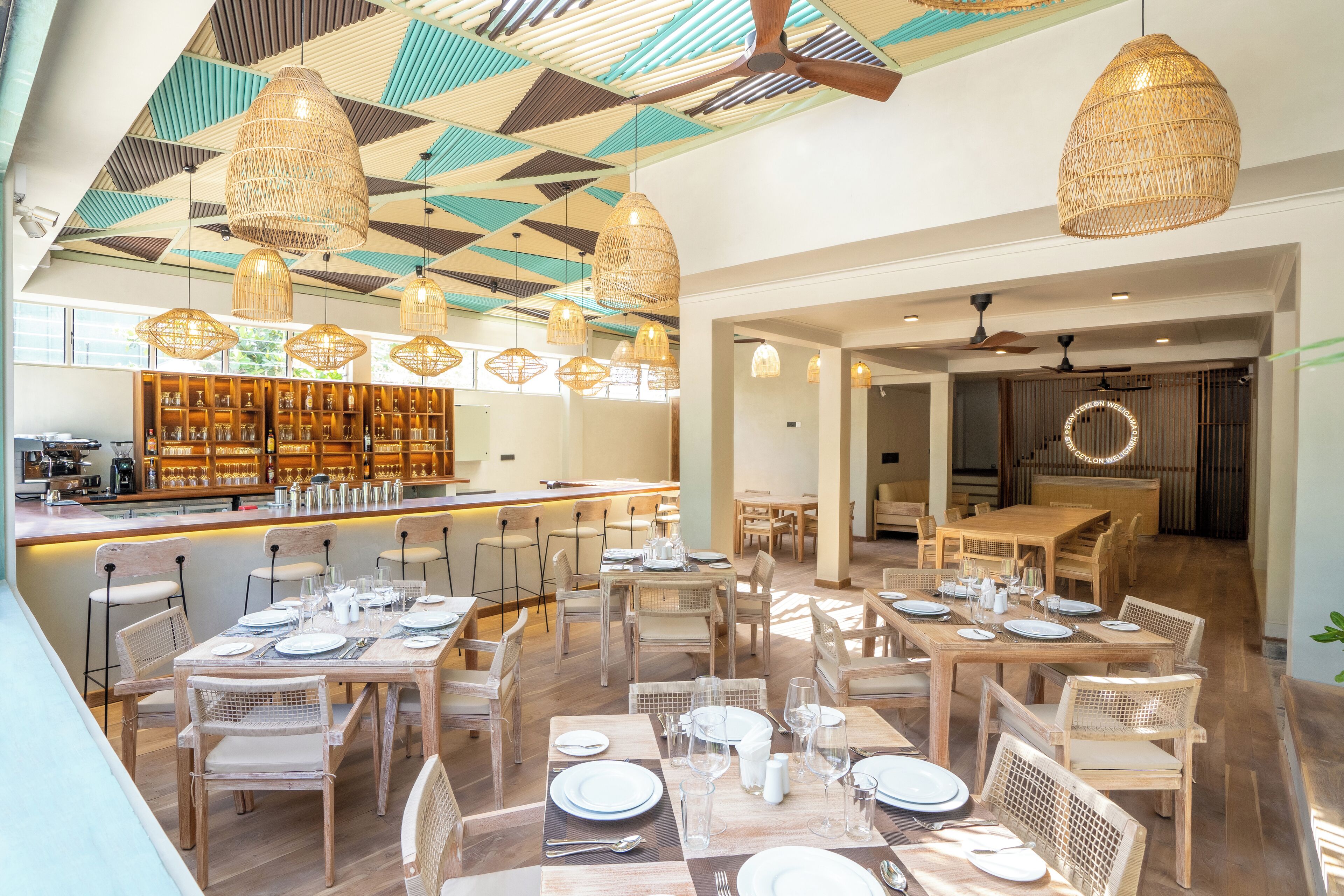 Uma sala de jantar ampla com várias mesas e cadeiras, um bar com prateleiras cheias de garrafas, e um teto com padrões geométricos.