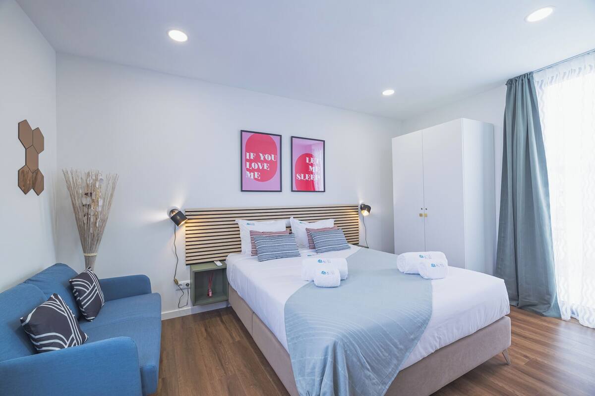 Quarto de hotel moderno com uma cama, um sofá azul e dois cartazes motivacionais na parede.