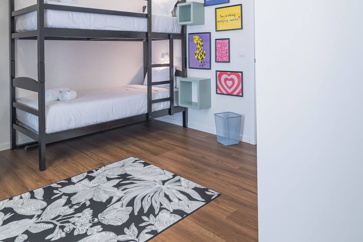 Quarto com beliche, tapete estampado com flores, uma lixeira pequena e várias fotos emolduradas na parede.