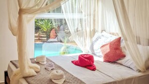 Uma cama com dossel, vista para uma piscina e planta tropical lá fora.