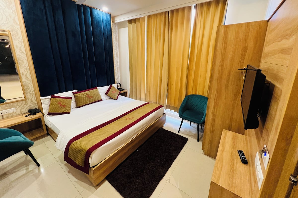 Quarto de hotel com uma cama, escrivaninha, cadeira, televisão e uma janela grande com cortinas.