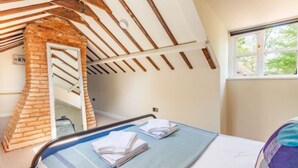 Um quarto com teto inclinado, uma chaminé de tijolos, uma cama com colchão azul e roupa de cama branca, e uma janela com vista para o verde.