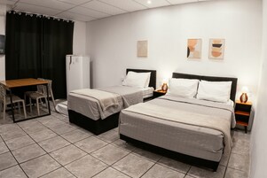 Quarto de hotel com duas camas, uma escrivaninha, cadeiras, um frigorífico e obras de arte nas paredes.