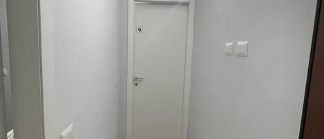 Um corredor estreito com uma porta branca identificada com o número 9, um interruptor de luz e uma câmera de segurança.