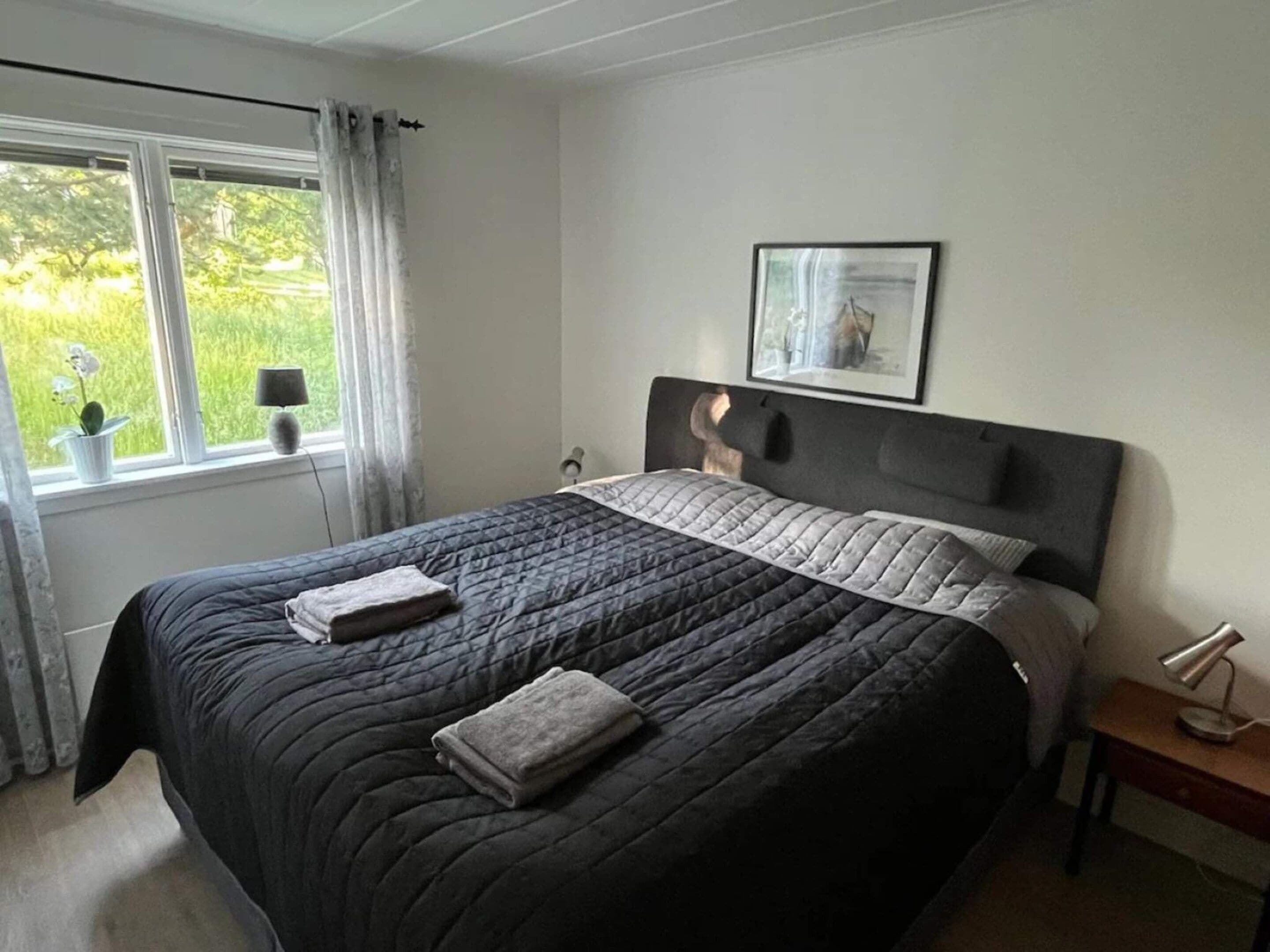 Quarto com cama, janela com cortinas, mesa de cabeceira com abajur e quadro na parede.