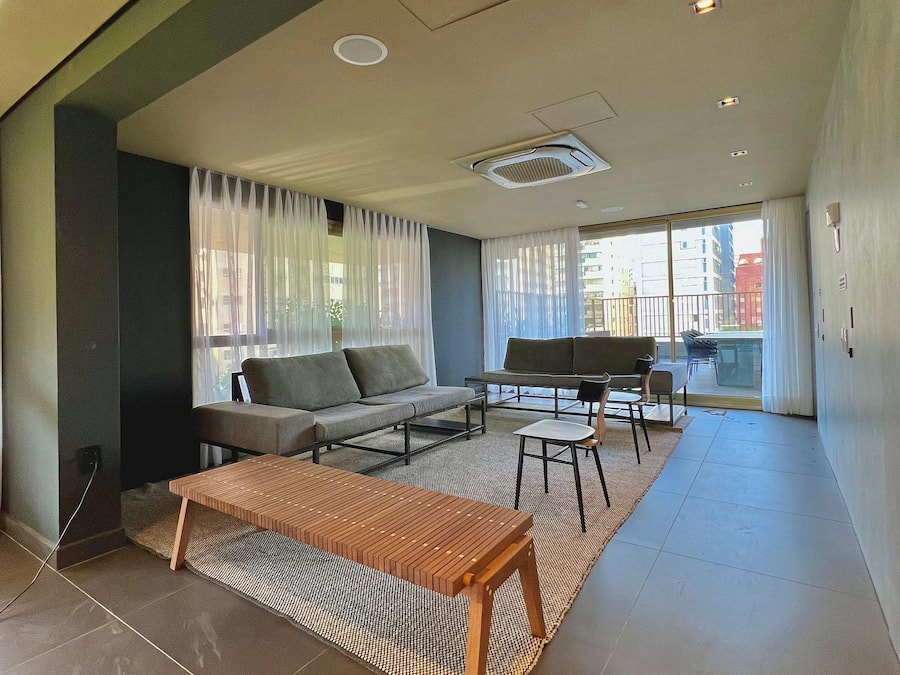 Uma sala de estar moderna com sofá, mesa de centro e cadeiras, uma janela grande com cortinas e vista para prédios.