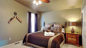 Quarto com cama, mesa de cabeceira, abajur, janela com cortinas e ventilador de teto.