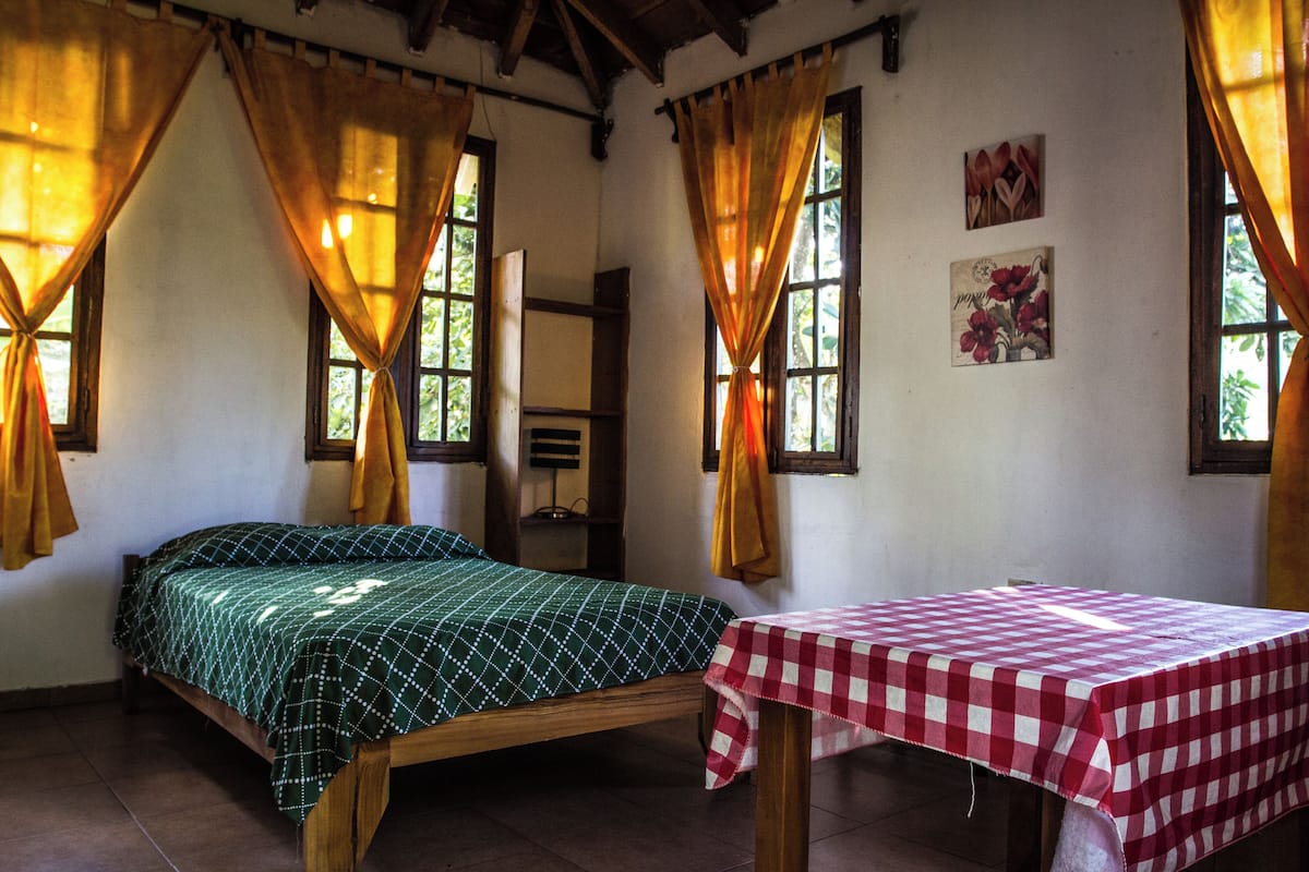 Um quarto com uma cama e uma mesa, ambas com toalhas de mesa xadrez, e cortinas nas janelas.