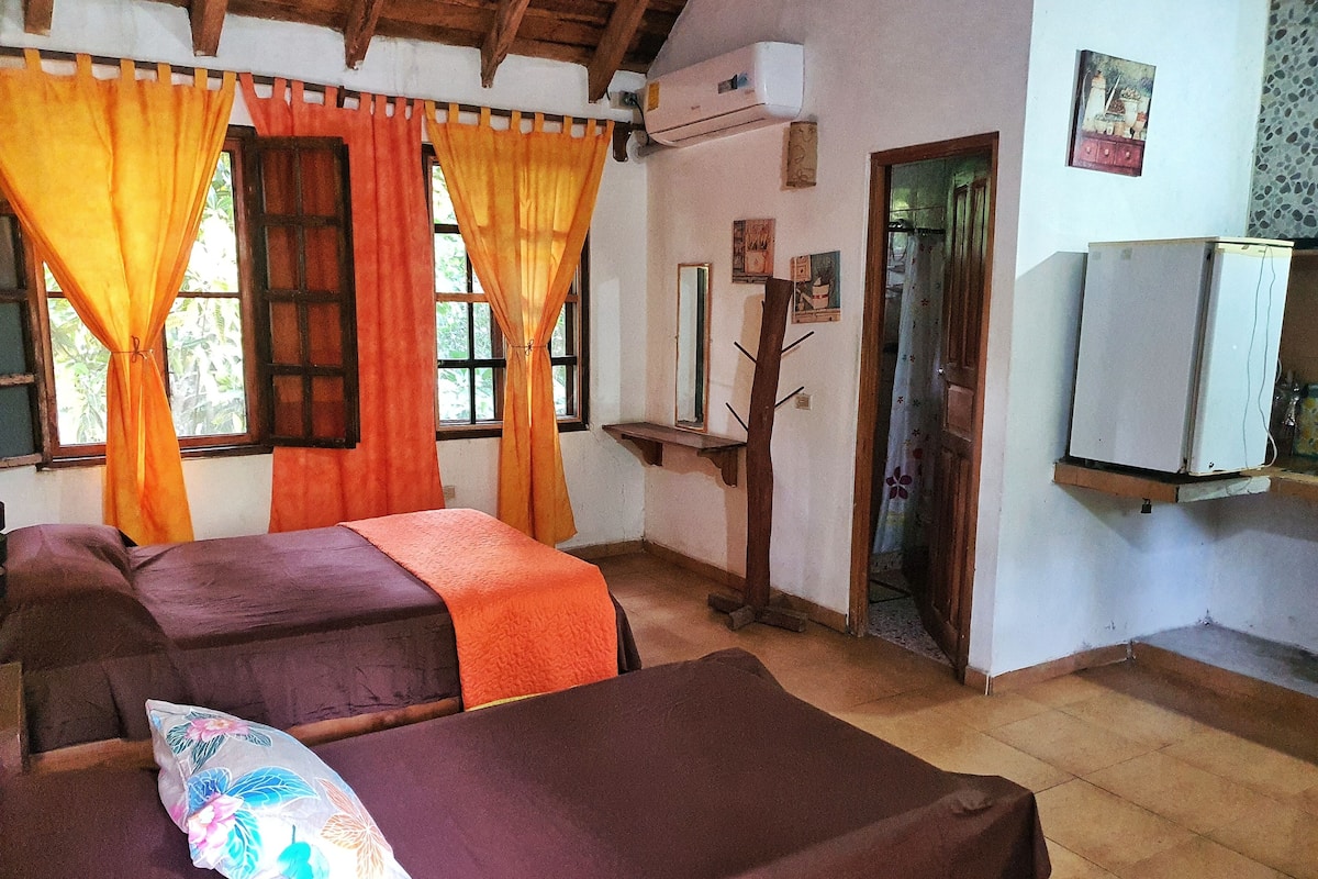 Quarto com duas camas, teto de madeira, ar-condicionado, uma prateleira pequena e porta que dá para outro cômodo.