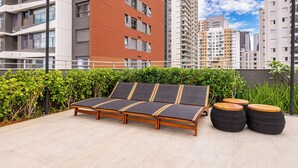 Uma área de estar moderna ao ar livre, com um sofá e duas mesas redondas, cercada por vegetação e com vista para uma paisagem urbana com edifícios altos.