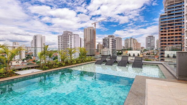 Piscina no terraço com espreguiçadeiras, rodeada por edifícios altos e um céu claro.