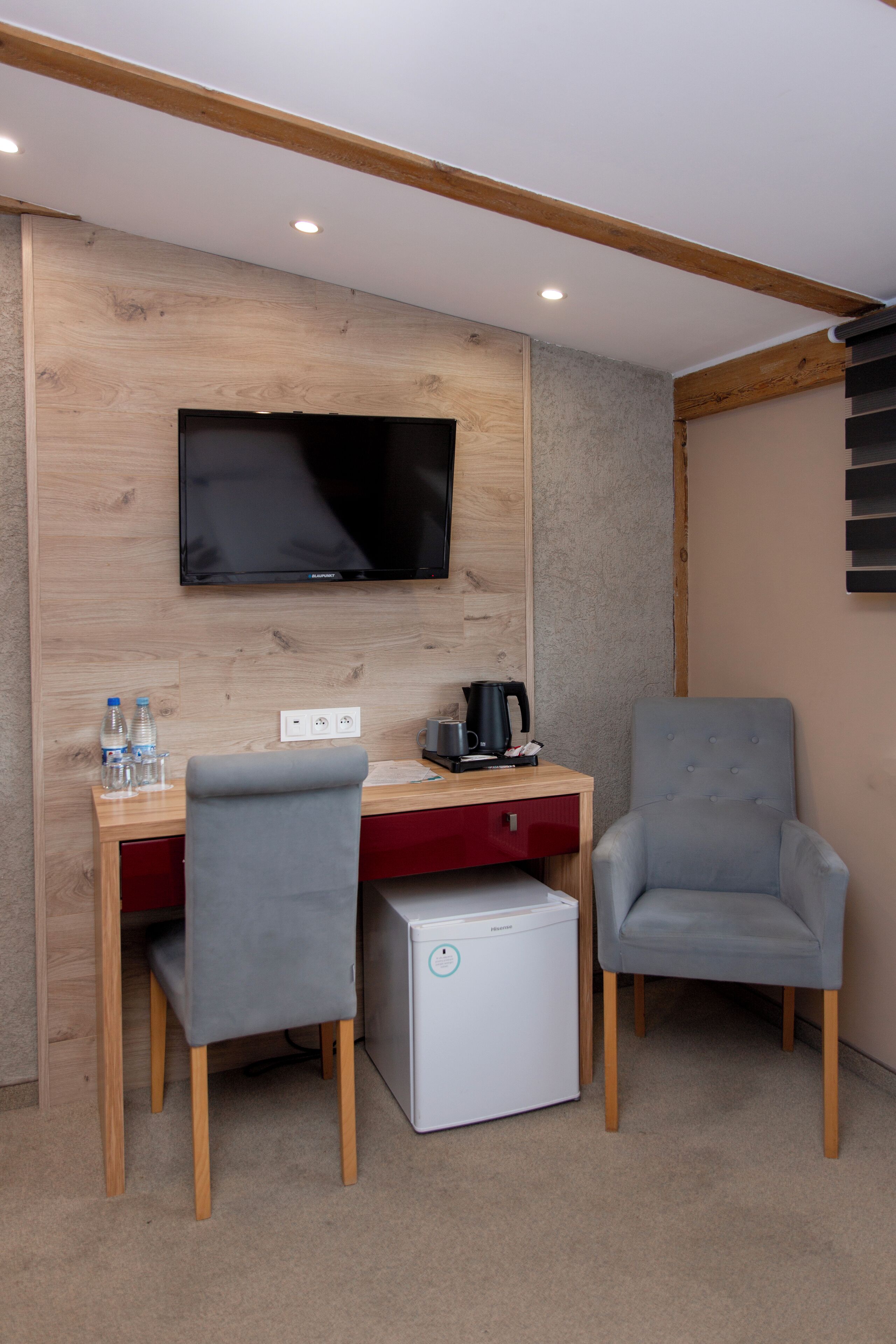 Quarto compacto com uma secretária de madeira, uma poltrona cinza, um frigorífico branco pequeno, uma TV de ecrã plano na parede e uma máquina de café.