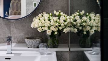 Uma casa de banho com uma pia branca, uma bacia de pedra, dois vasos de vidro transparente com flores brancas e um espelho refletindo as flores e a pia.