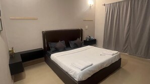 Cama bem arrumada com edredom branco e travesseiros, mesa de cabeceira com abajur e ventilador de teto.