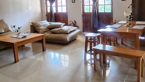 Uma sala de estar ampla com sofá, mesa de centro de madeira e mesa de jantar com bancos. Há janelas grandes com cortinas, um aparelho de ar condicionado e um ventilador de teto.