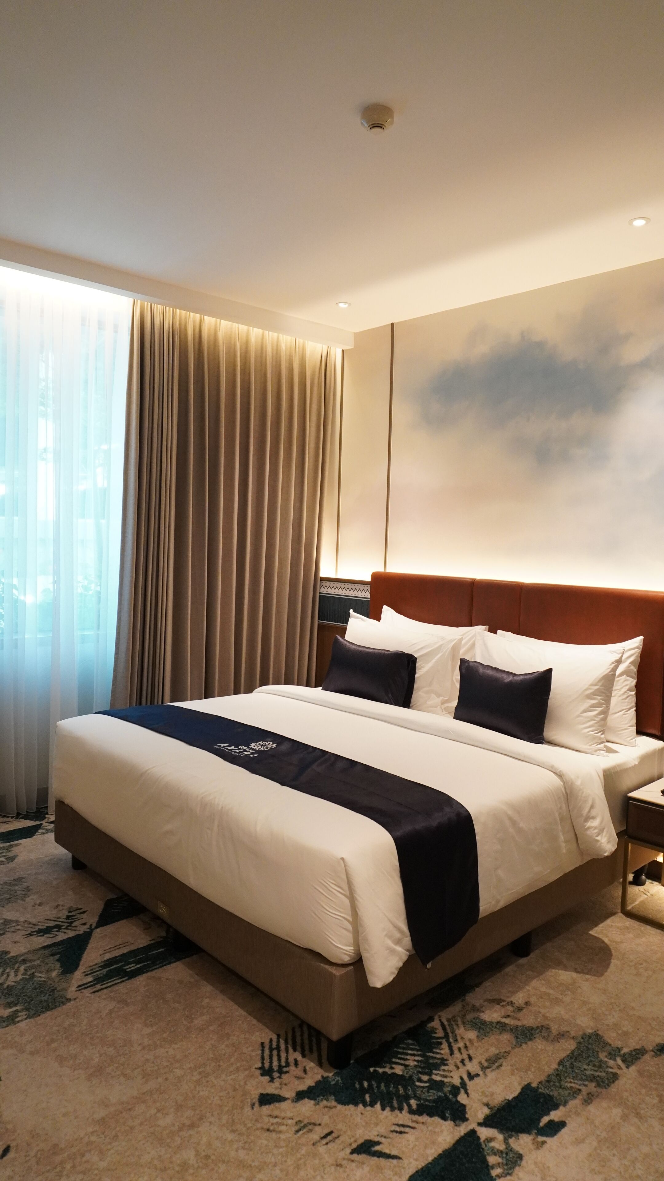 Quarto de hotel com uma cama grande, mesa de cabeceira, luminária na parede e janela com cortinas.