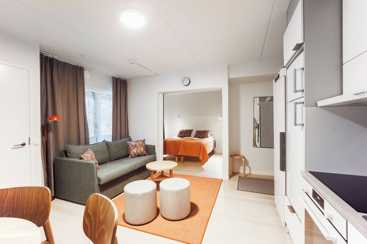 Quarto de hotel moderno com sofá, dois pufes, mesa de jantar, cama, televisão e cozinha compacta.